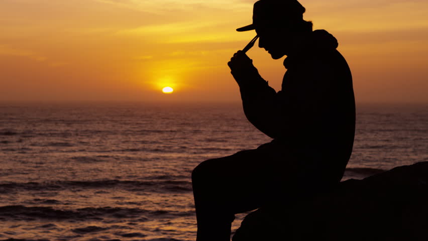 Young Man Putting On Sunglasses And Staring Out At Sunset Over Ocean ...